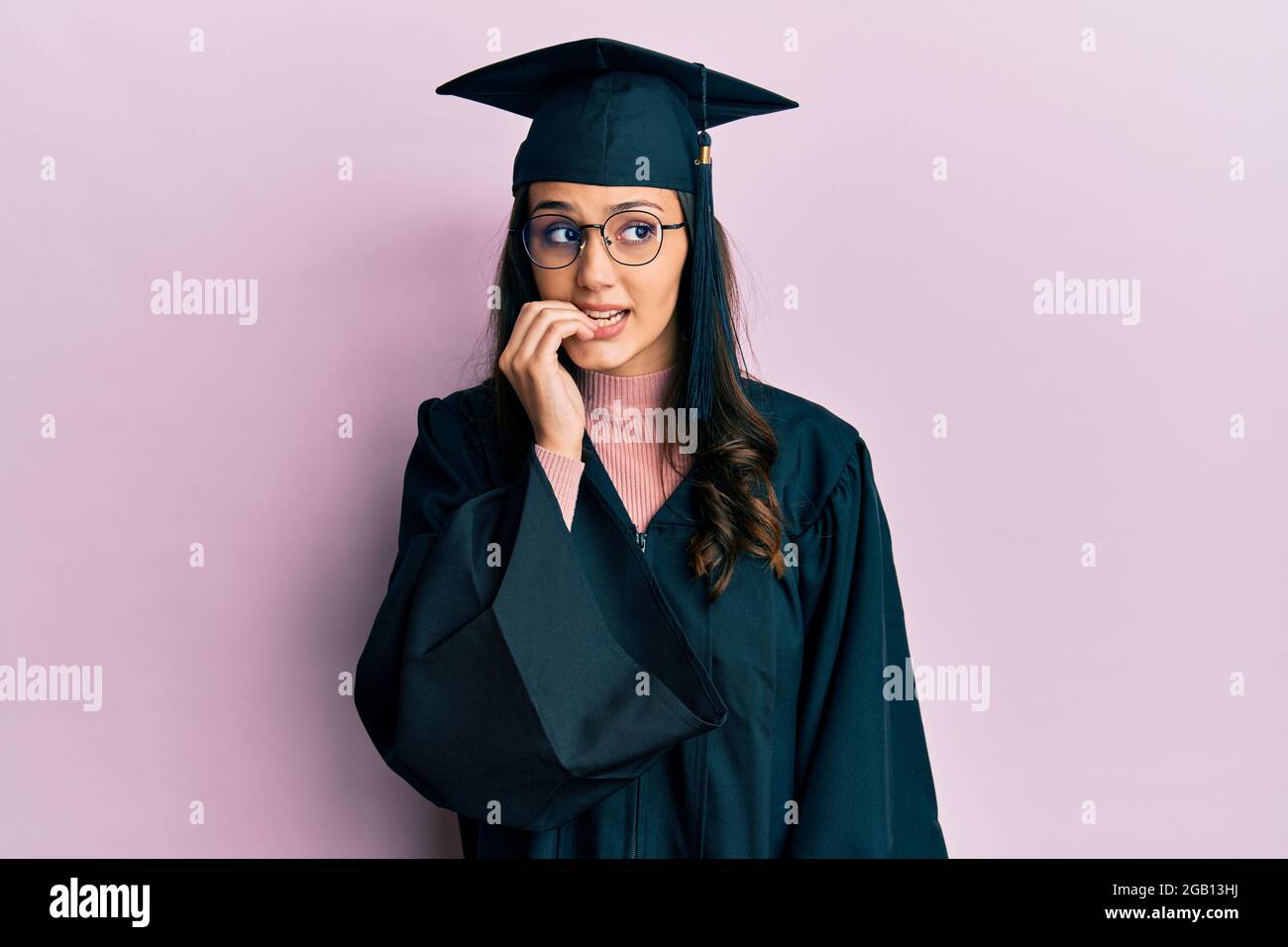 Young hispanic woman wearing graduation cap and ceremony robe looking ...