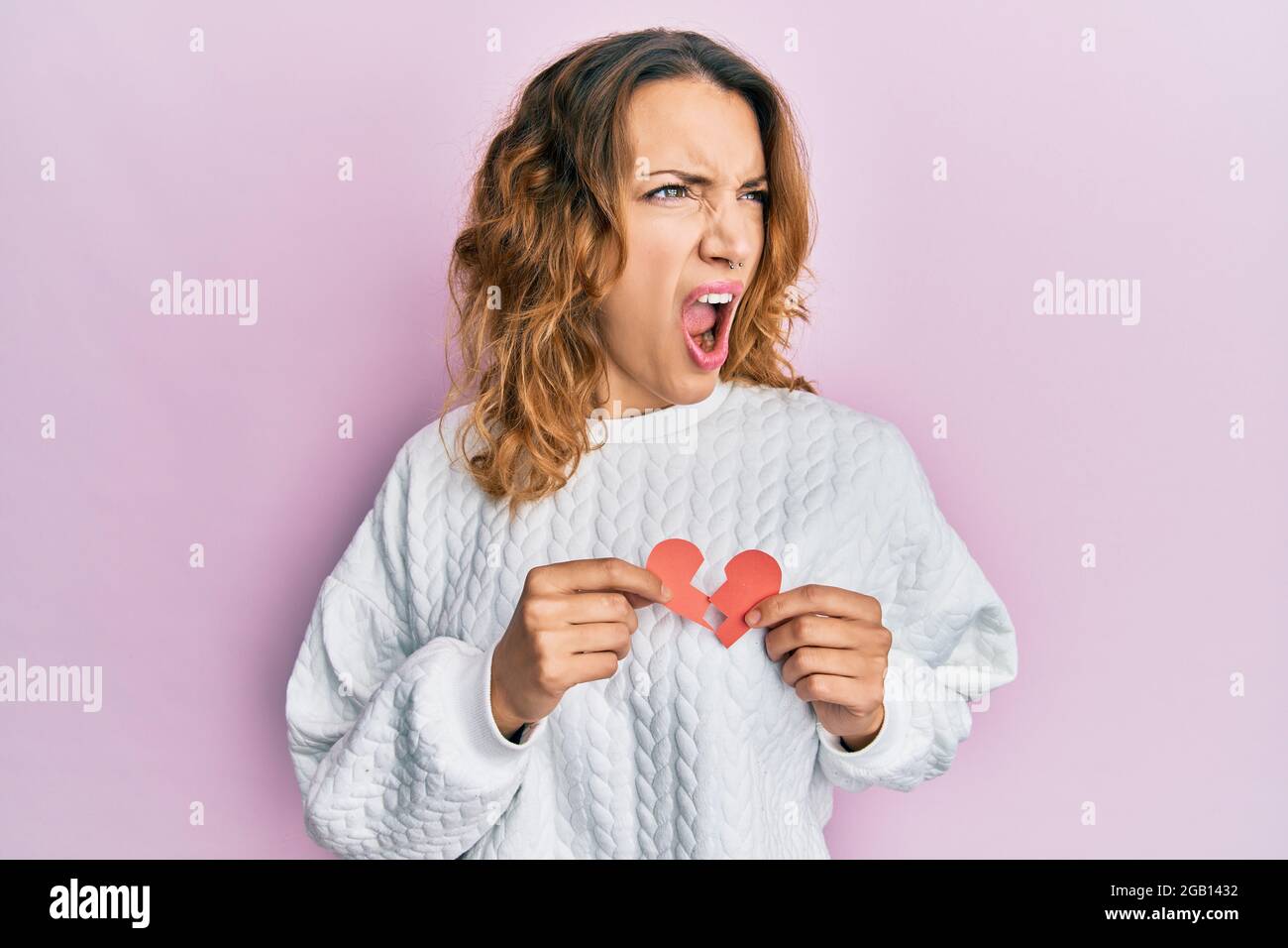Young caucasian woman holding broken heart paper shape angry and mad ...