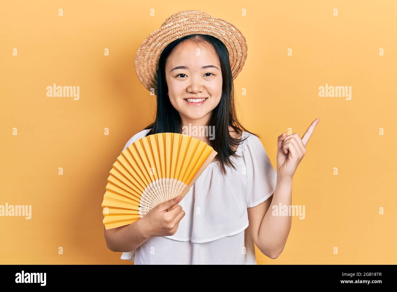 Young chinese girl waving hand fan cooling air in summer smiling happy pointing with hand and ...