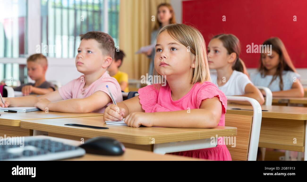 Interested blonde tween girl listening to schoolteacher and writing in ...