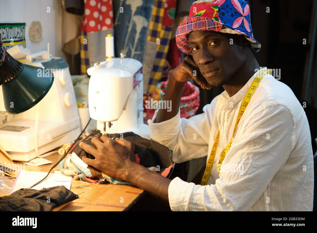 African tailor sewing a hat with a sewing machine looking at camera ...