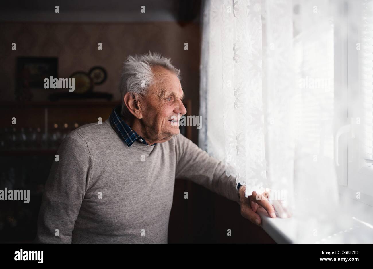 Portrait of elderly man standing indoors at home, looking out through ...