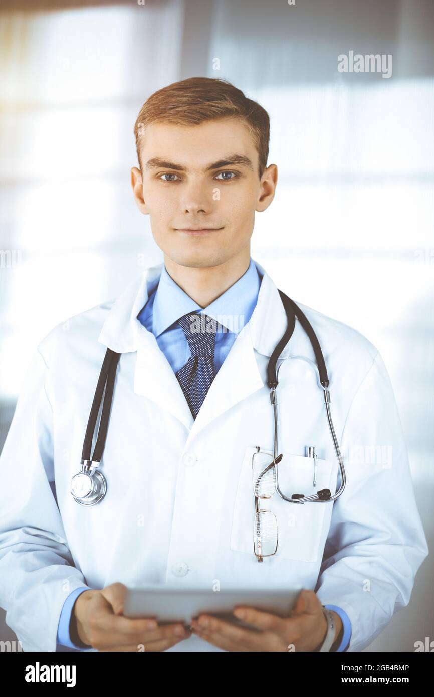 Young doctor man is smiling cheerfully at camera, while standing in a ...