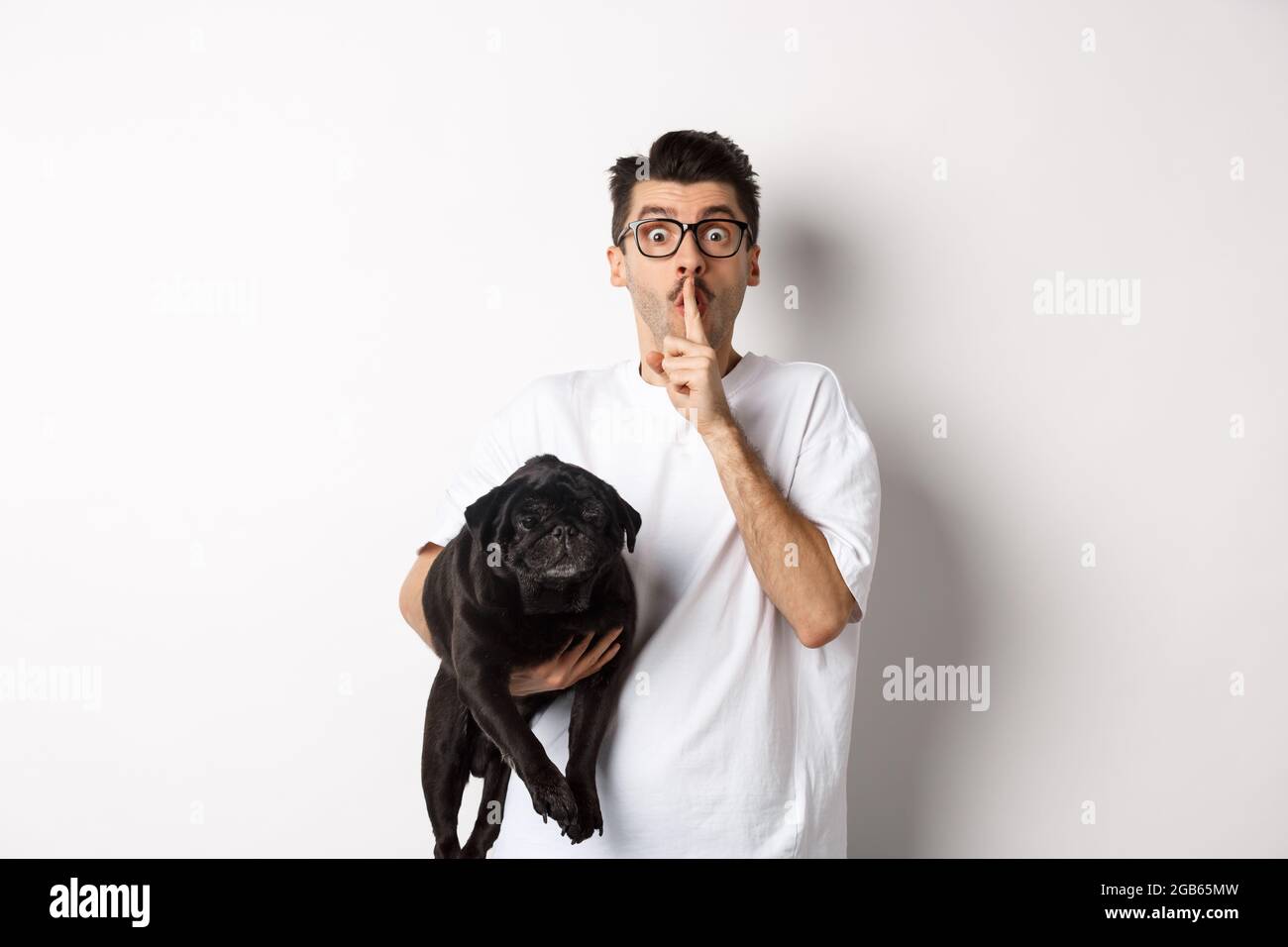 Image of funny young man holding cute black pug dog and shushing at ...