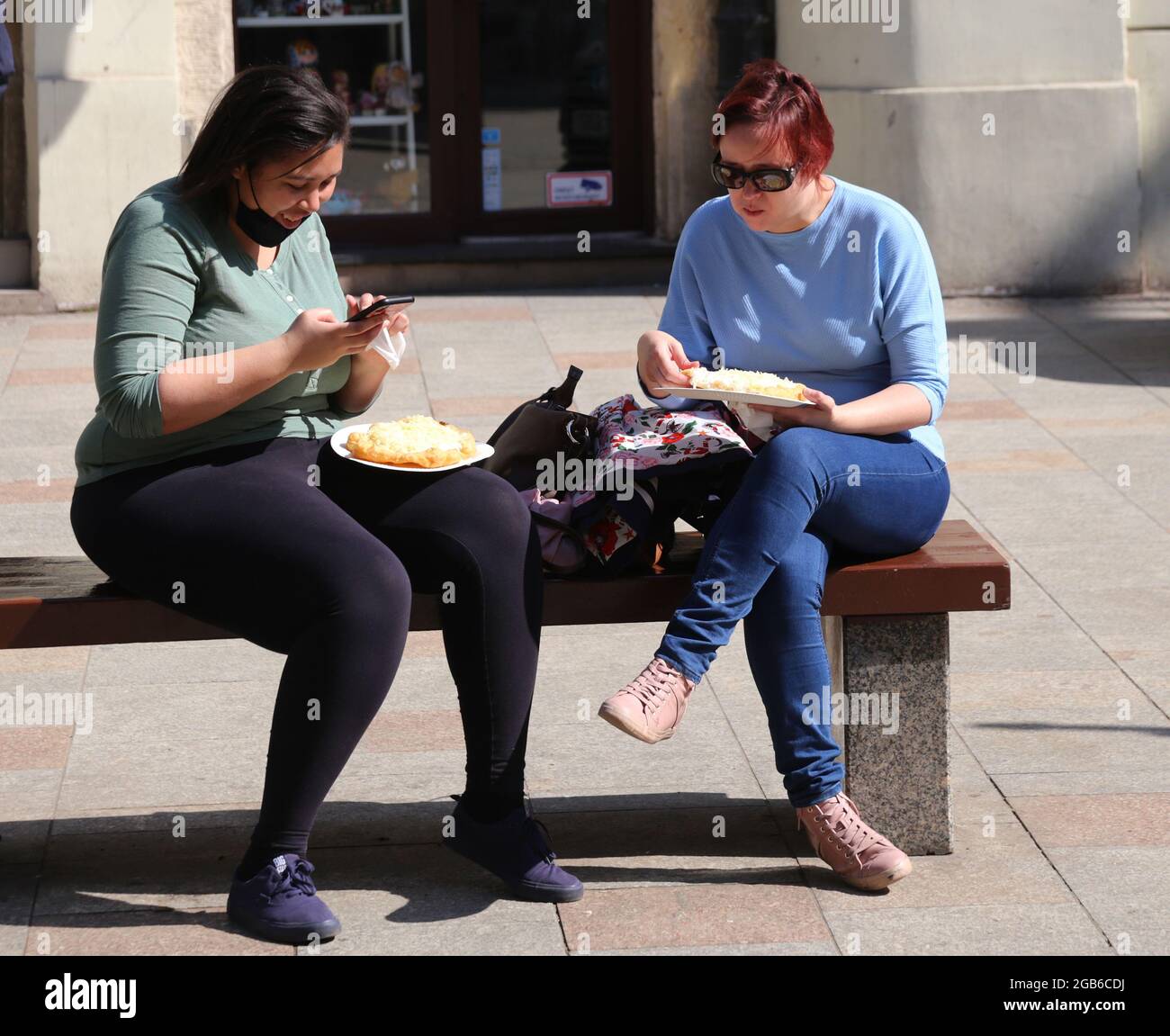 Two young women eating their pizzas sitting on the street bench in ...