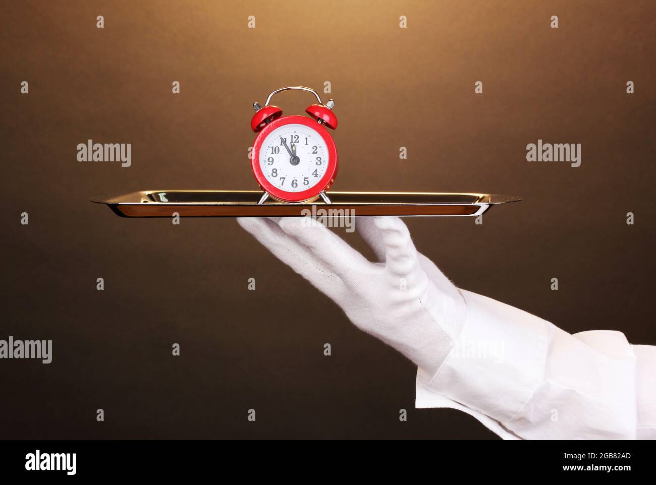 Hand in glove holding silver tray with alarm clock on brown background ...