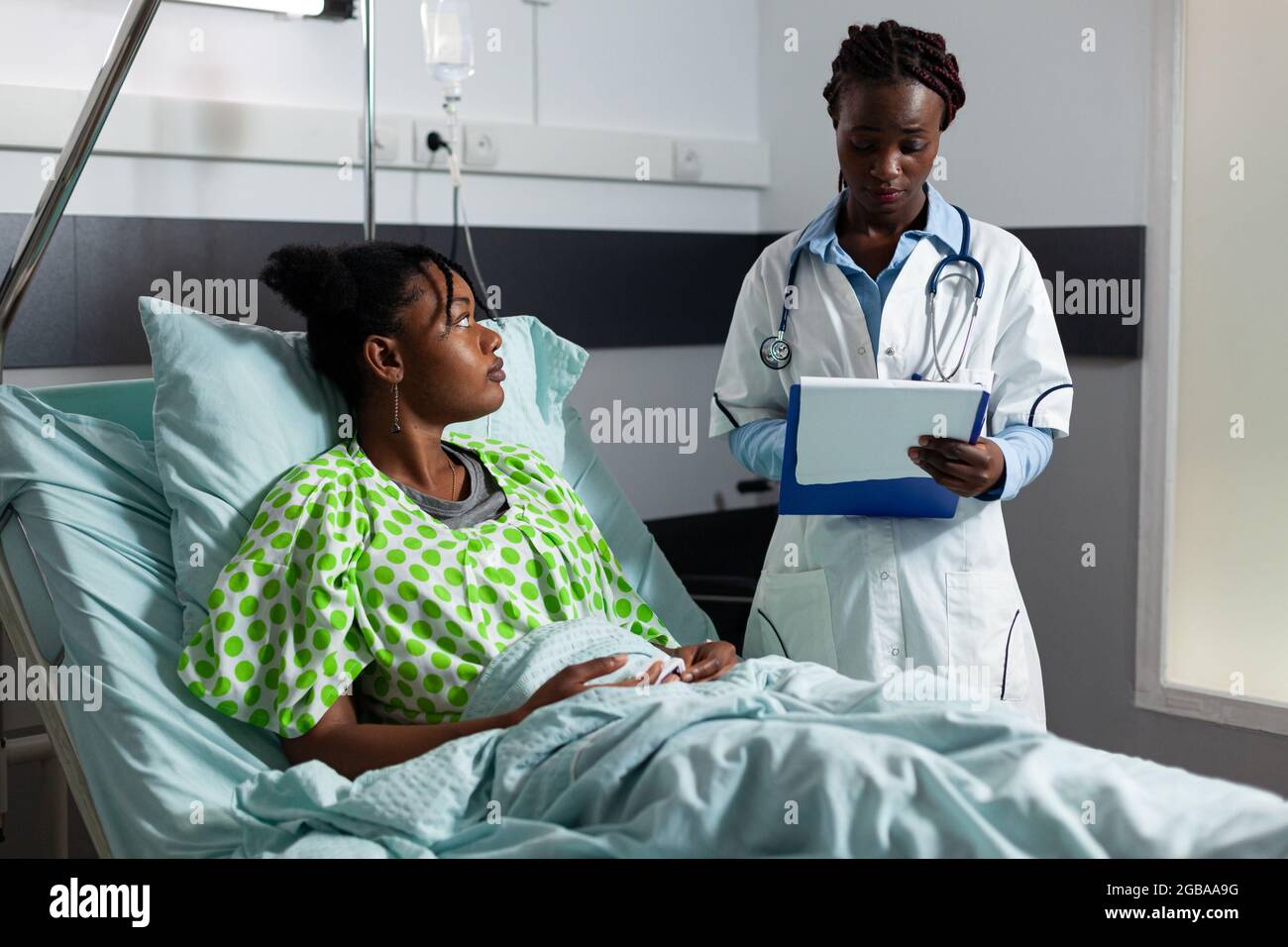 Professional Doctor Talking To Afro Patient Laying In Hospital Ward Bed