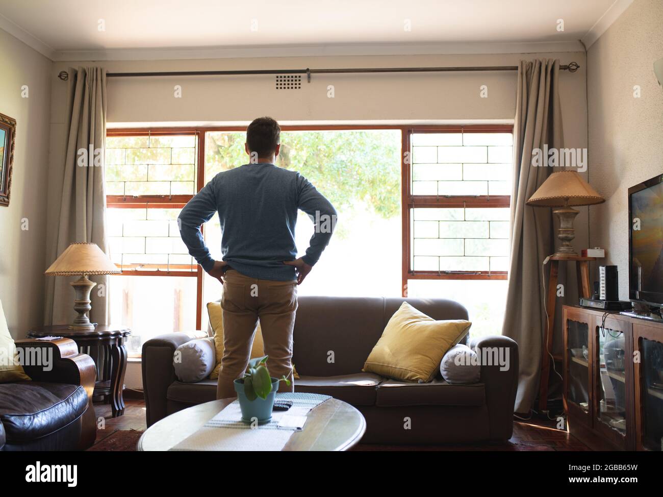 Back view of caucasian man standing in living room, looking out window ...