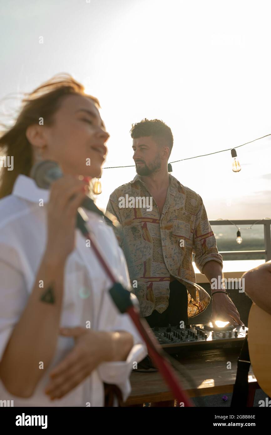Young man standing by soundboard during rooftop party with singing girl ...
