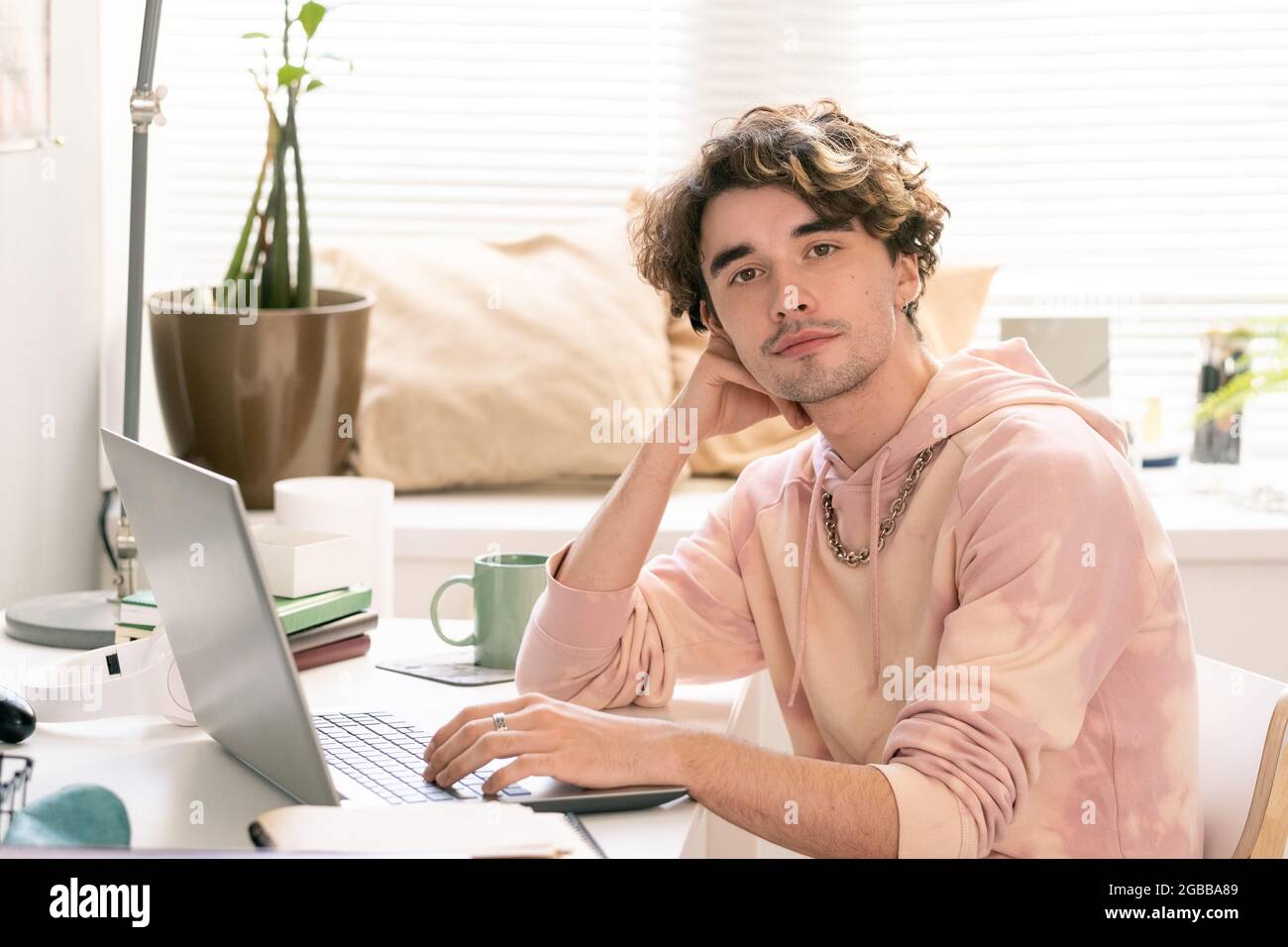 Handsome teenager in pink hoodie networking while sitting by desk in ...