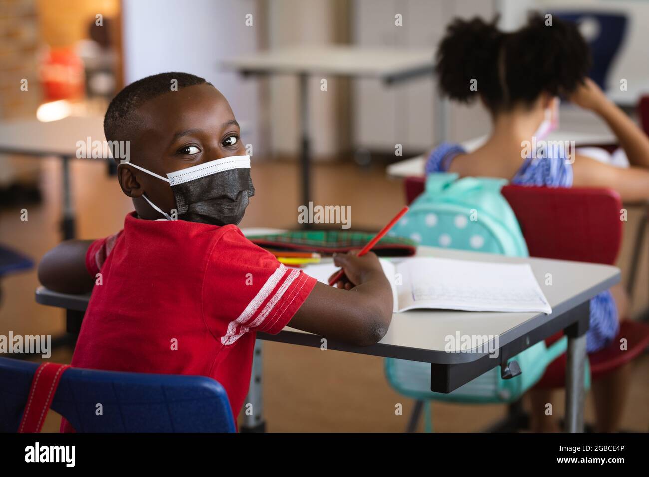 Portrait of african american boy wearing face mask sitting on his desk ...