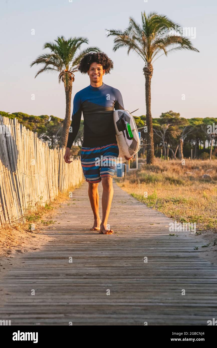 Young afro latino surfer walks down the wooden walkway to the beach ...