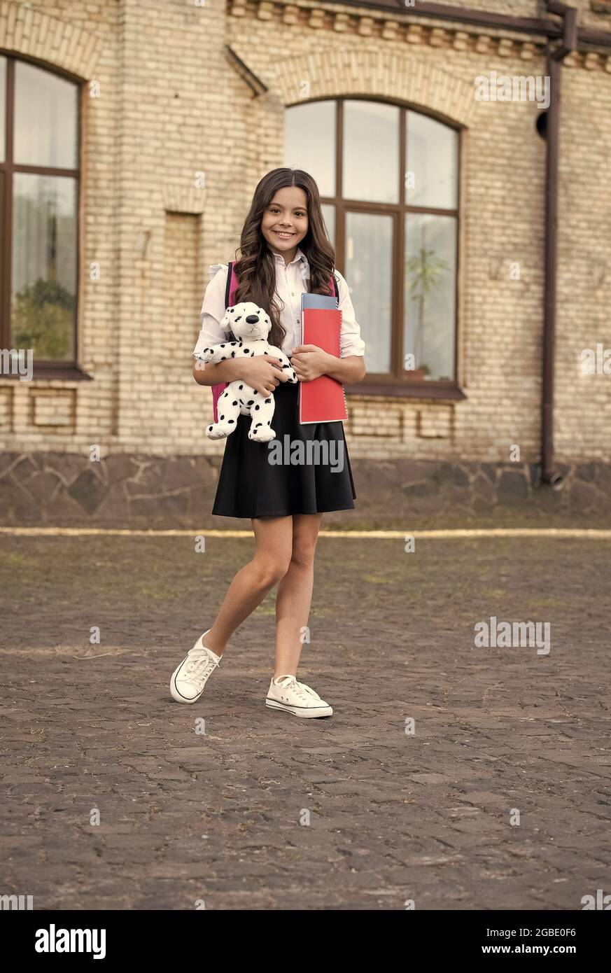 Happy small kid in school uniform hold books and toy dog in schoolyard ...