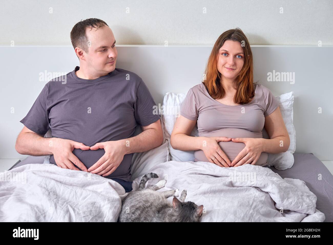 Pregnant wife and overweight husband lie in bed holding their bellies ...