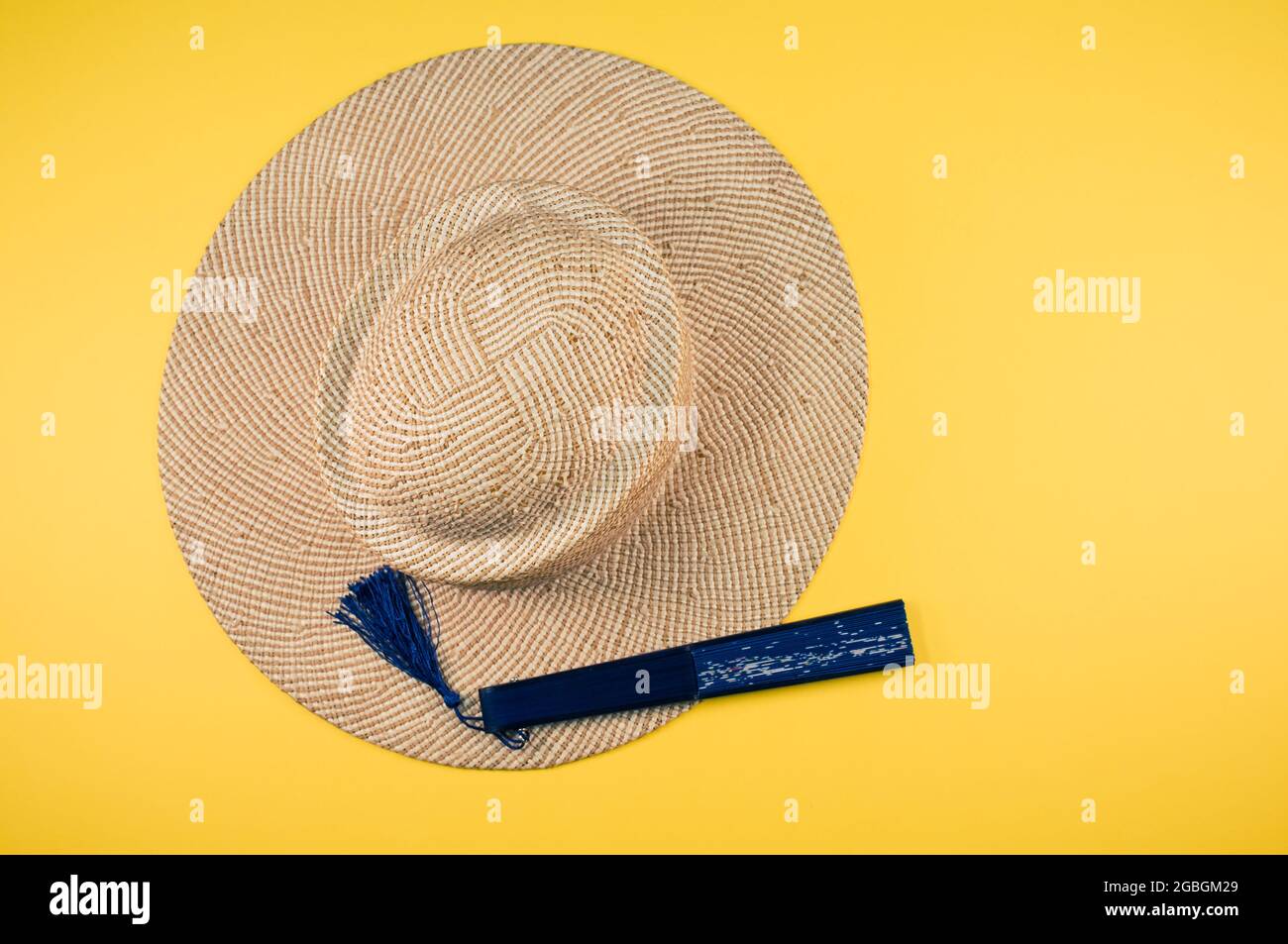 Round straw hat and a blue hand fan isolated on a yellow background ...