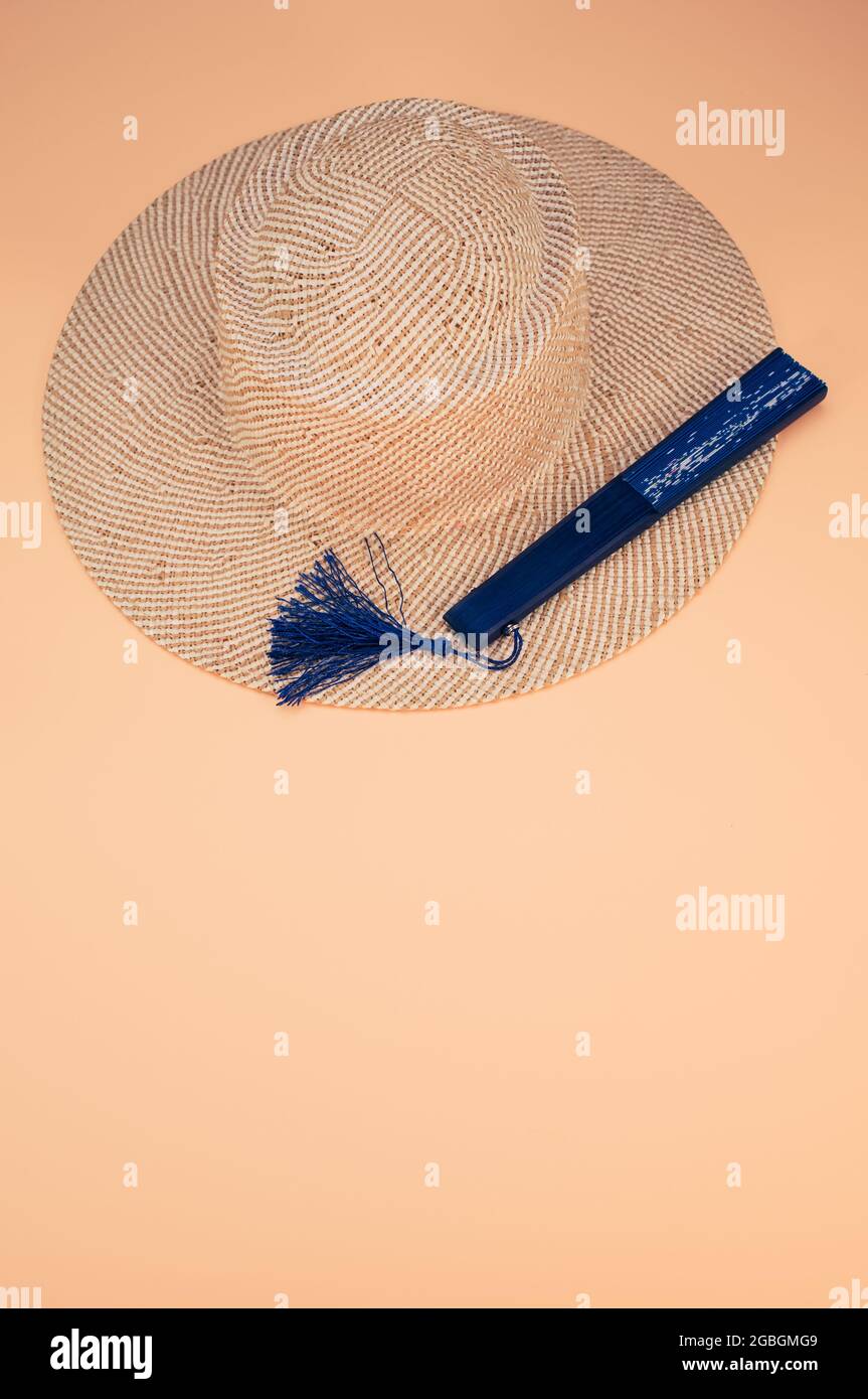 Round straw hat and a blue hand fan isolated on a peach background with ...