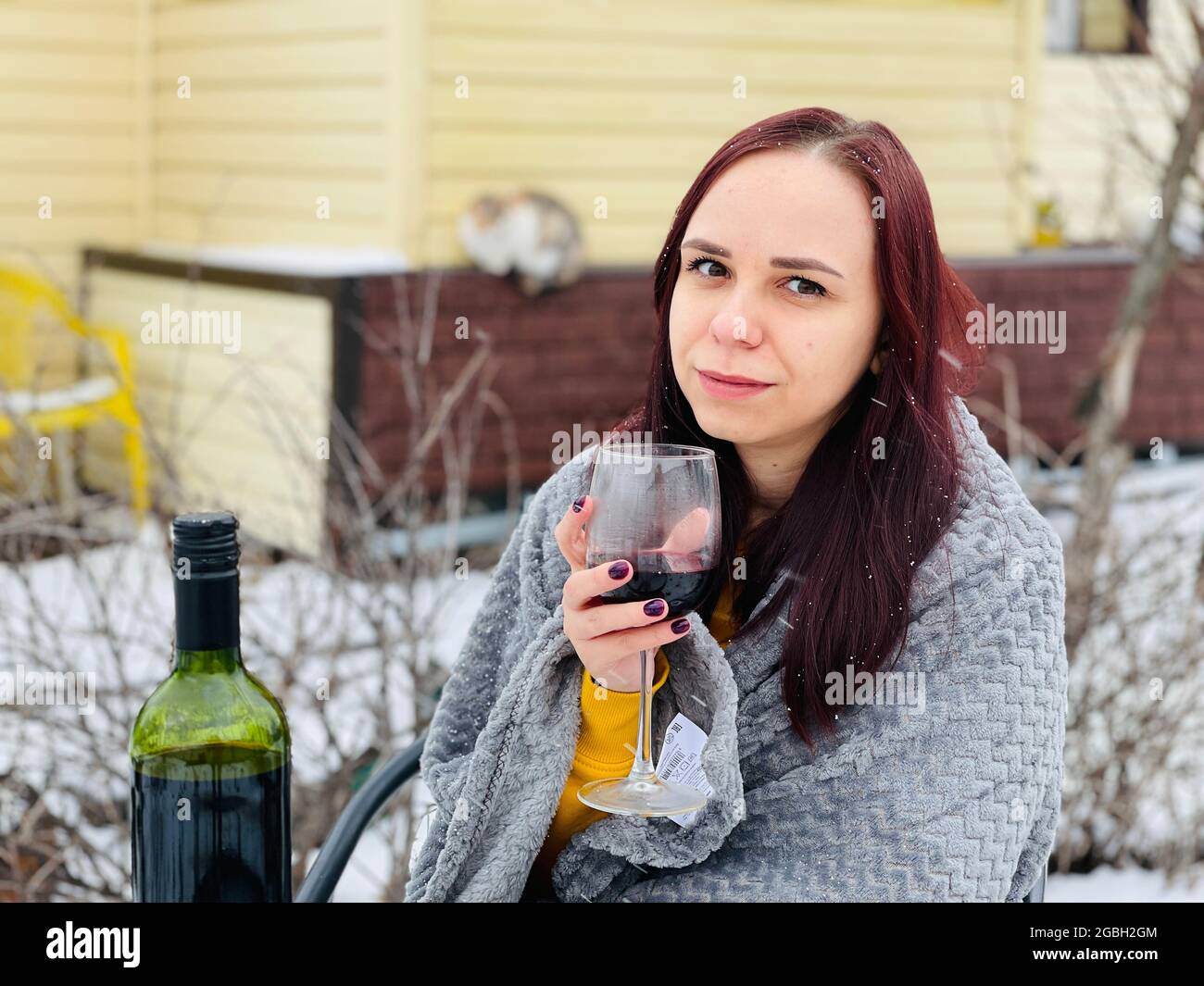 Young woman sitting in yard and drinking red wine in snowy weather ...