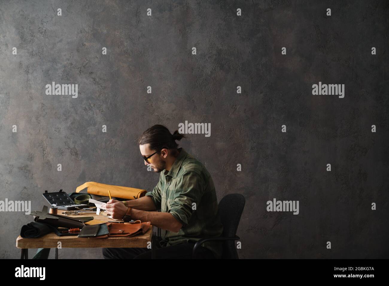 Bearded focused craftsman writing down notes while working with leather ...