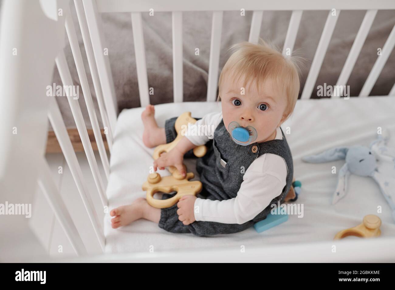 Portrait of serious baby boy with dummy sitting in bed and playing with ...