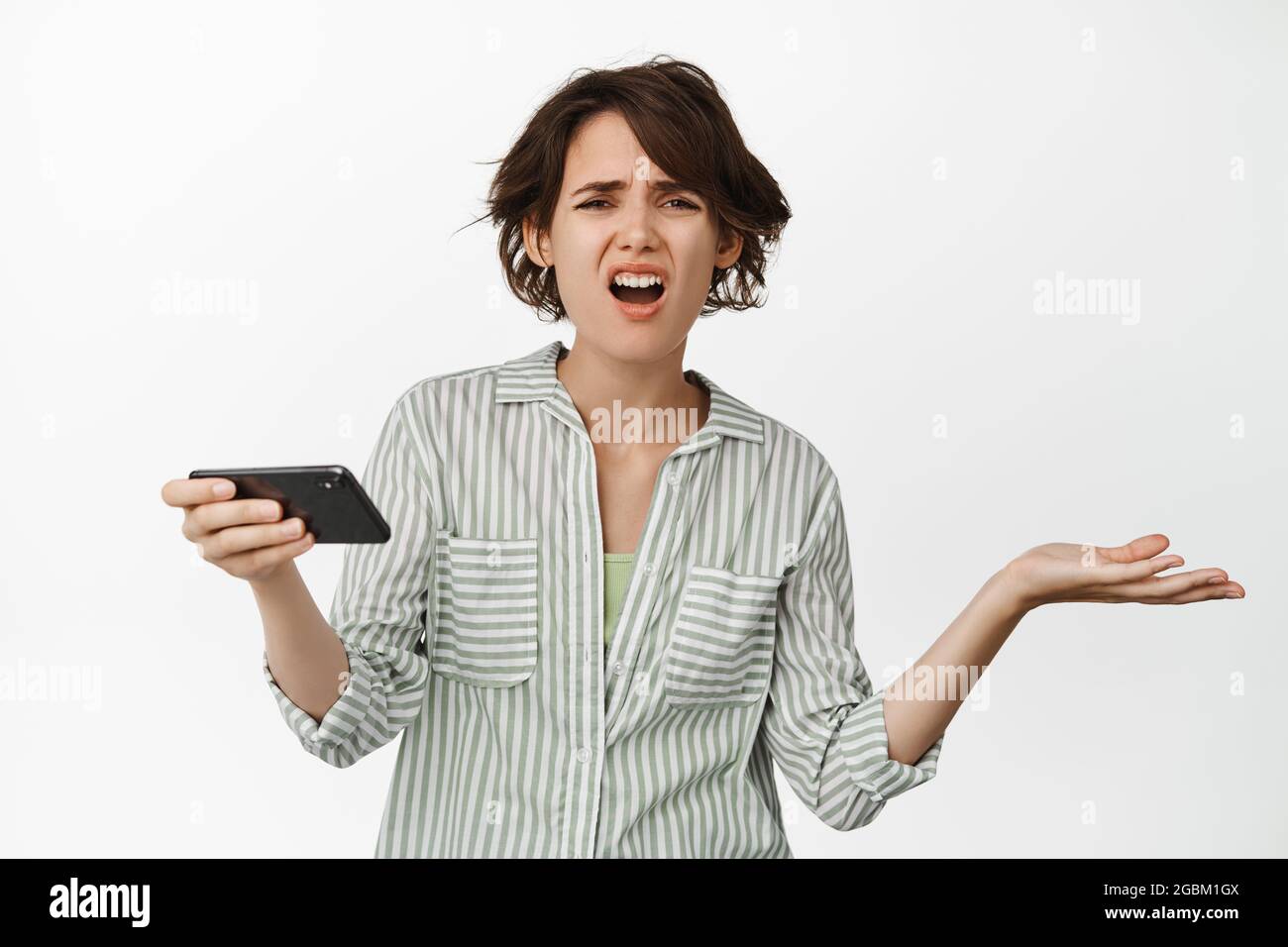 Image of frustrated girl holding mobile phone and shrugging, frowning puzzled, cant understand ...