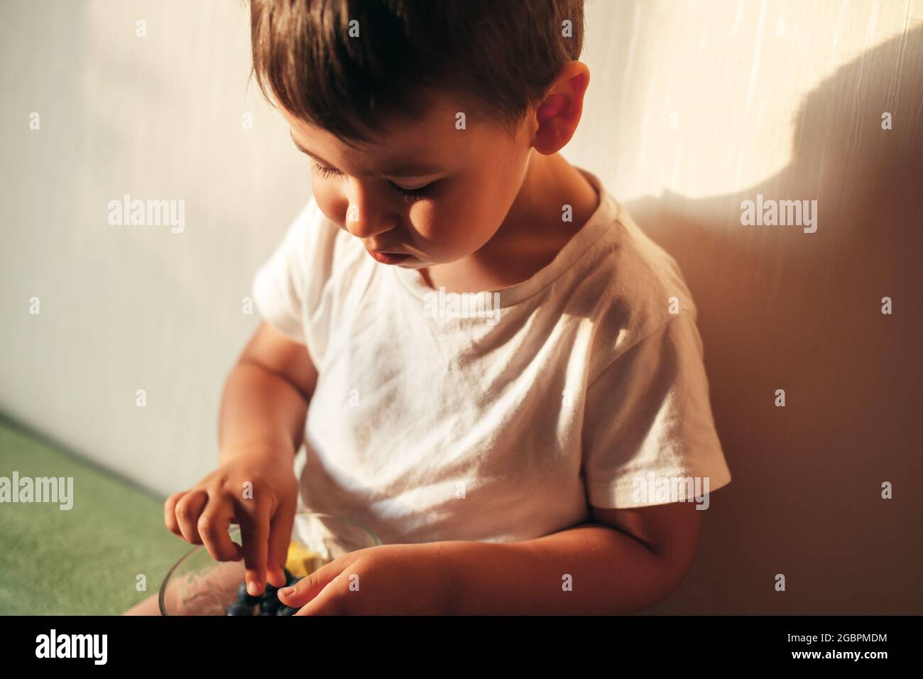 Adorable caucasian boy is eating blueberries at home wearing a white ...