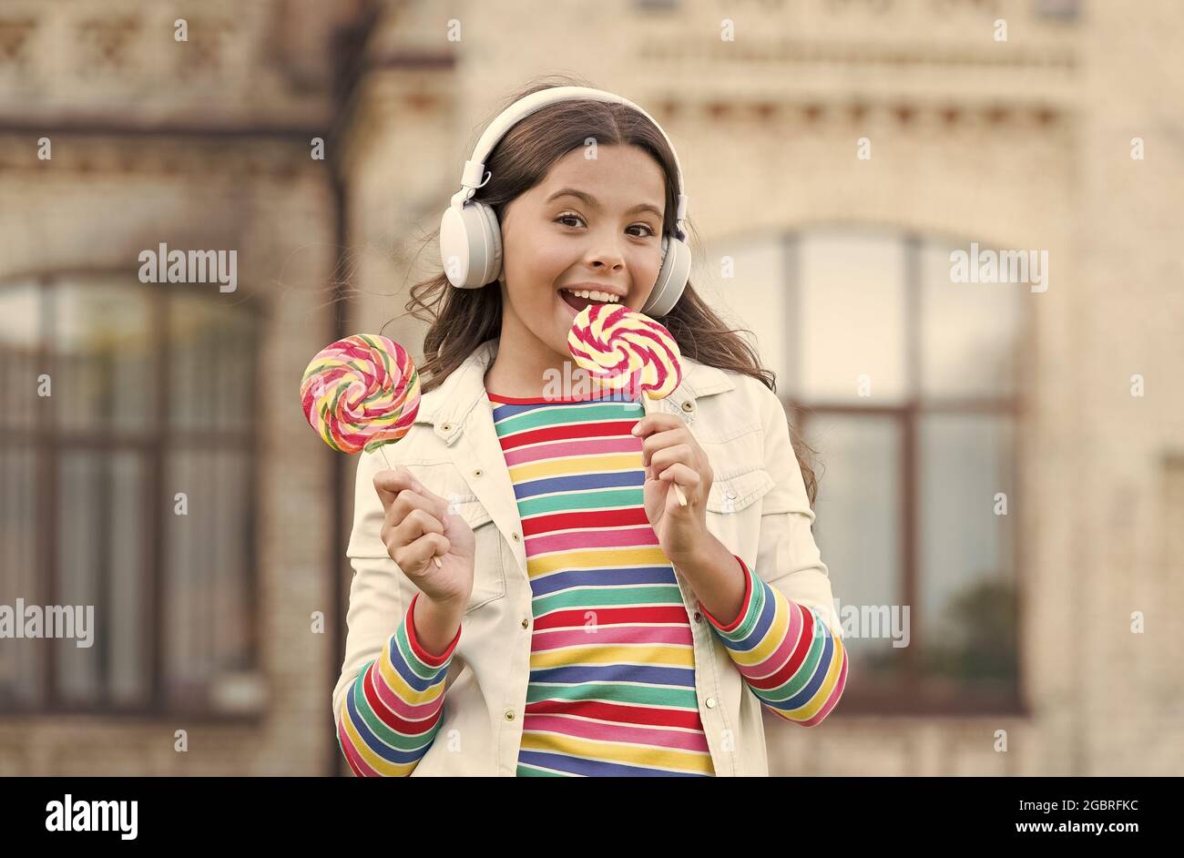 Cheerful girl modern wireless headphones eat candies, yummy lollipop ...
