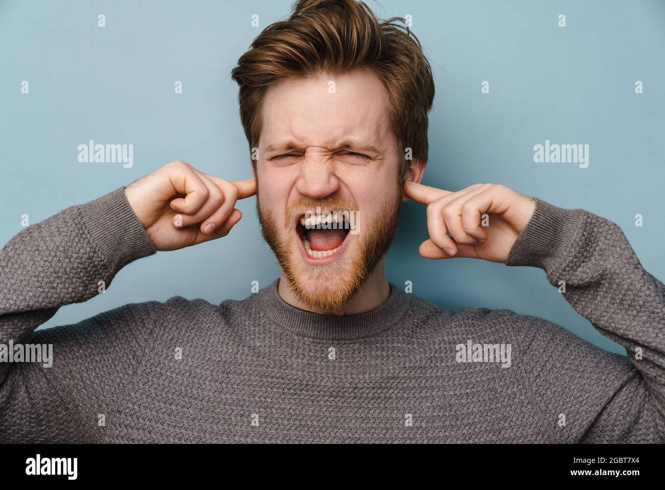 Bearded ginger man screaming while plugging his ears isolated over blue ...