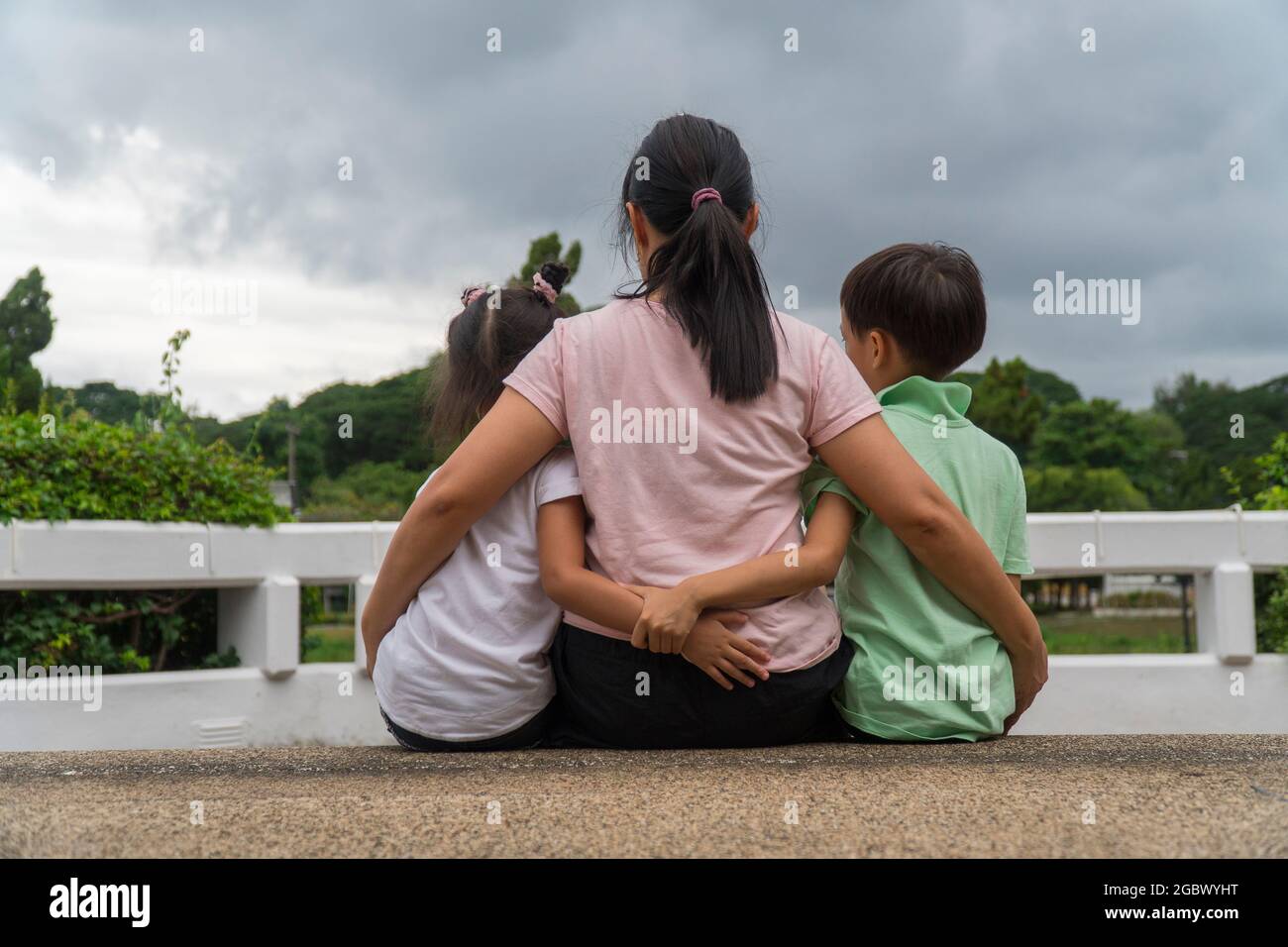 Back view of loving Asian mother hug her kids sitting on bridge, caring ...