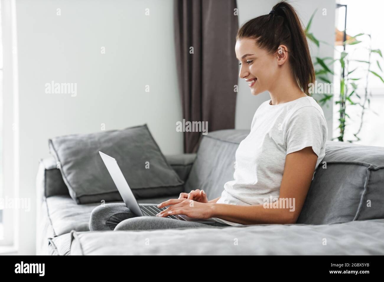 European smiling woman using laptop while sitting on couch at home ...
