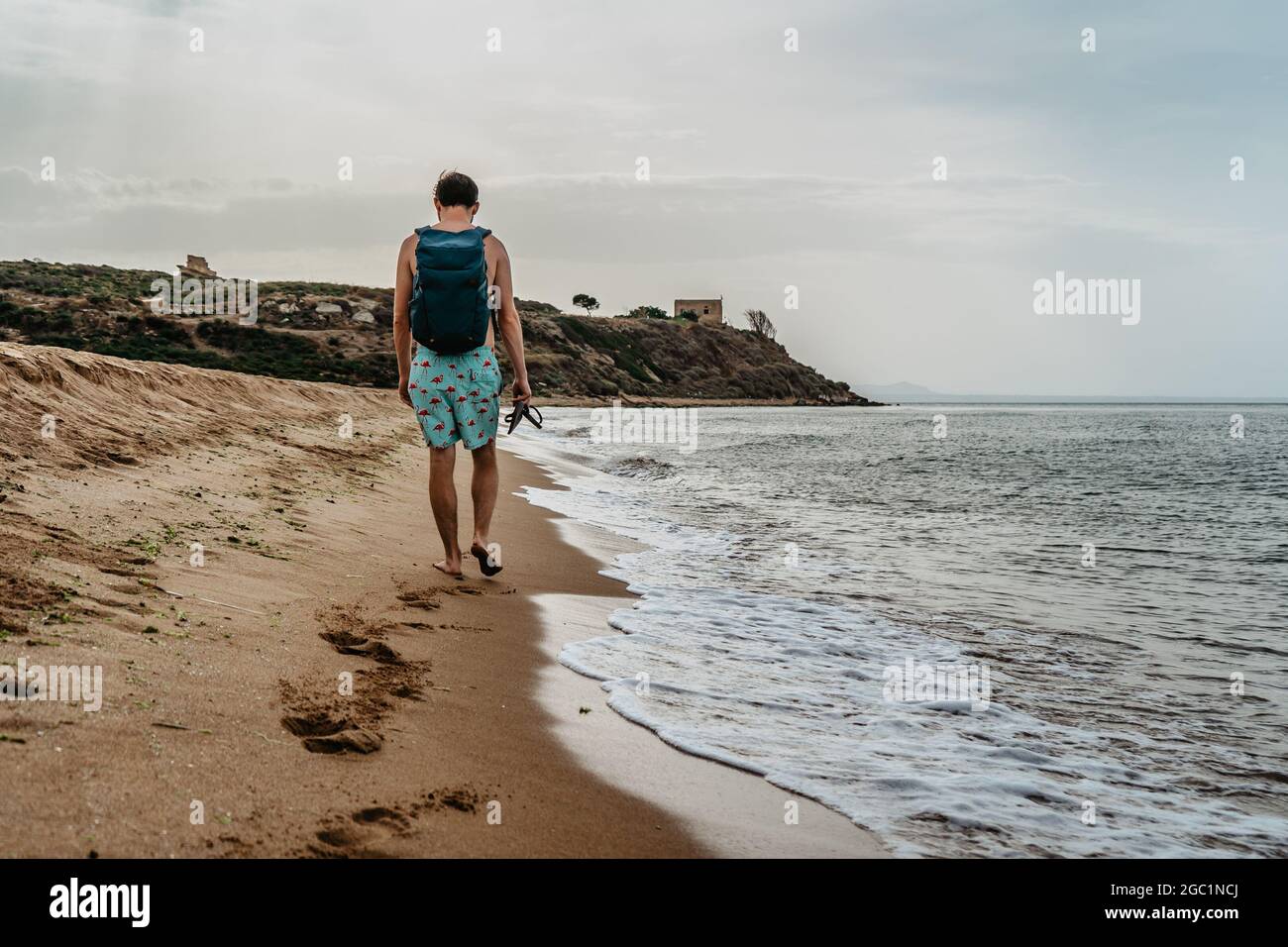 Shirtless man with backpack walking barefoot on empty sandy beach back ...