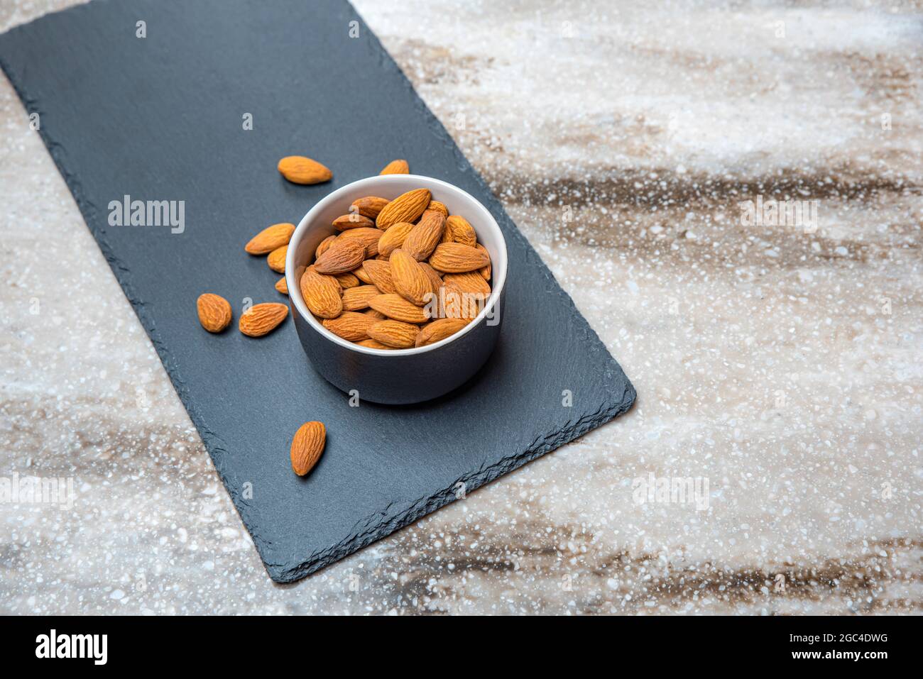 Almonds in a saucer on a black stone table top. Almonds are scattered ...