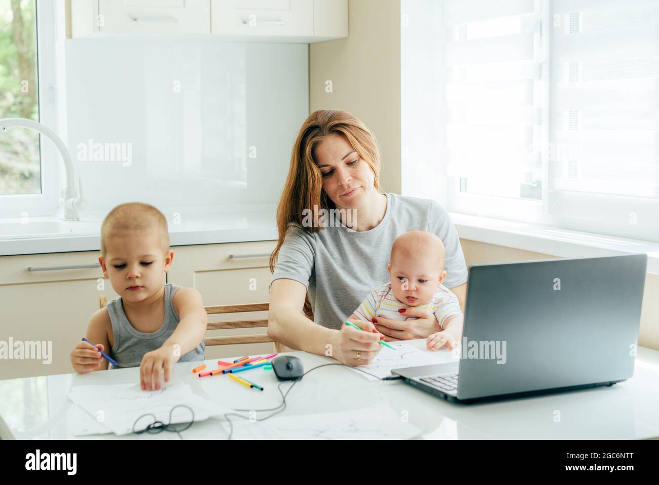 Young tired mom works on a laptop and babysits children in the kitchen ...