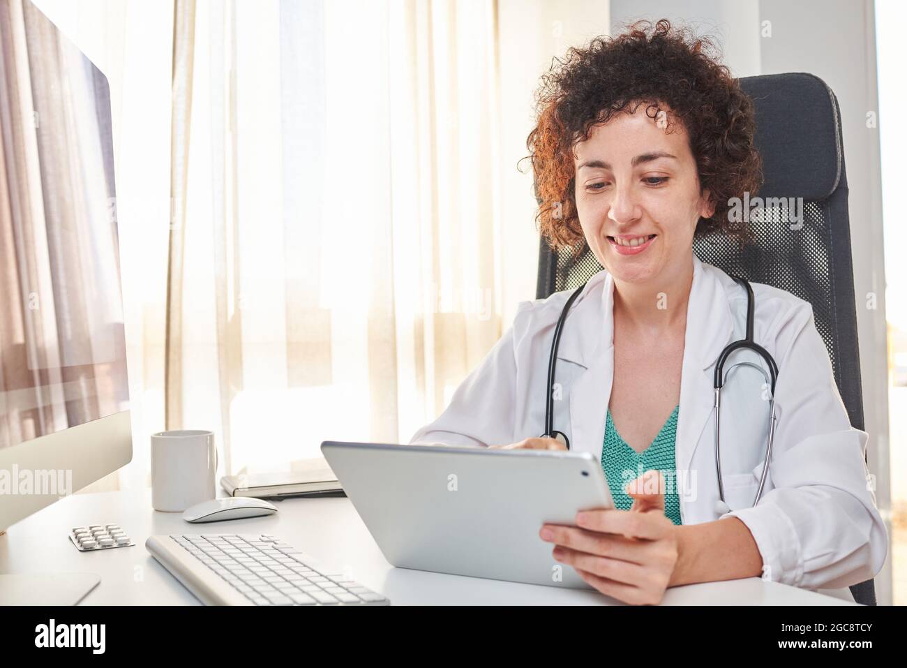 A female doctor uses her digital tablet sitting at the desk in her ...