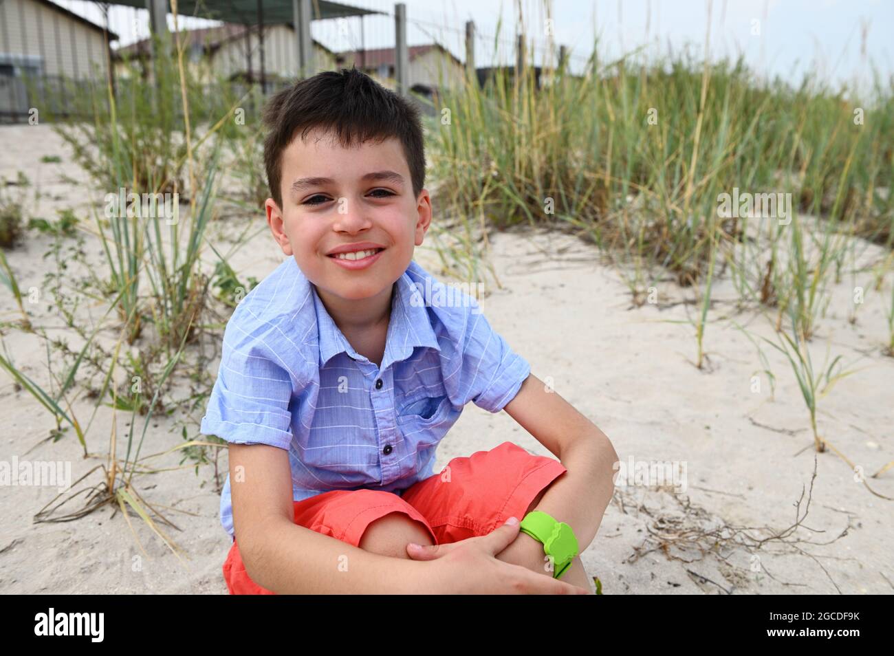 A handsome schoolboy sits on the sandy ground in a lotus position and ...