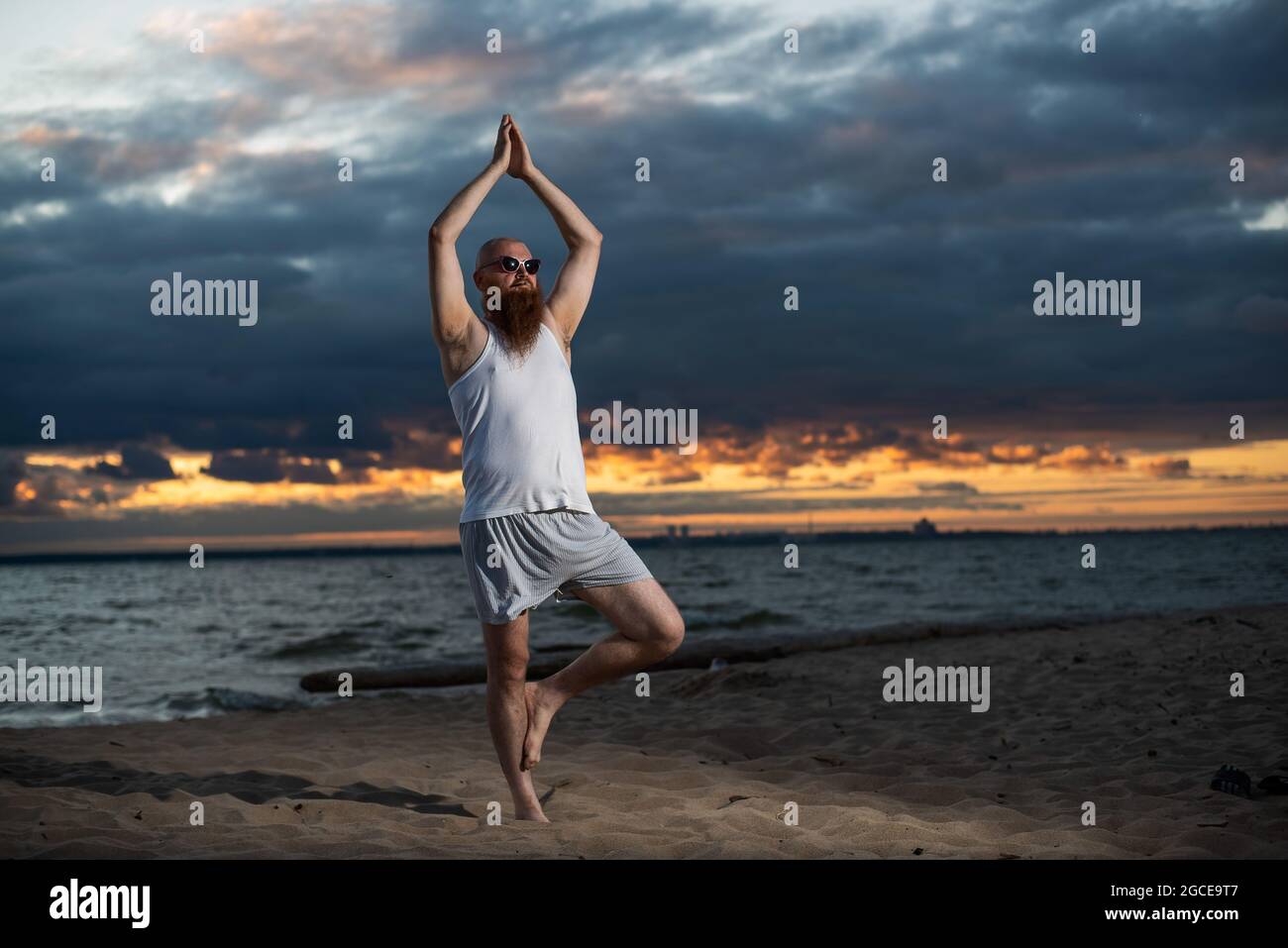 A bald man with a red beard practices yoga on the beach at sunset. A ...