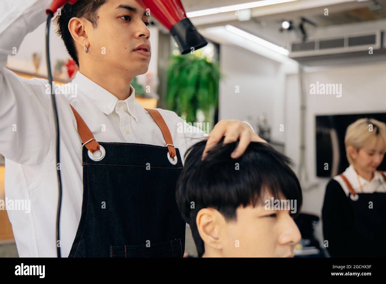 Young male Asian hairdresser wearing apron drying handsome young man's ...
