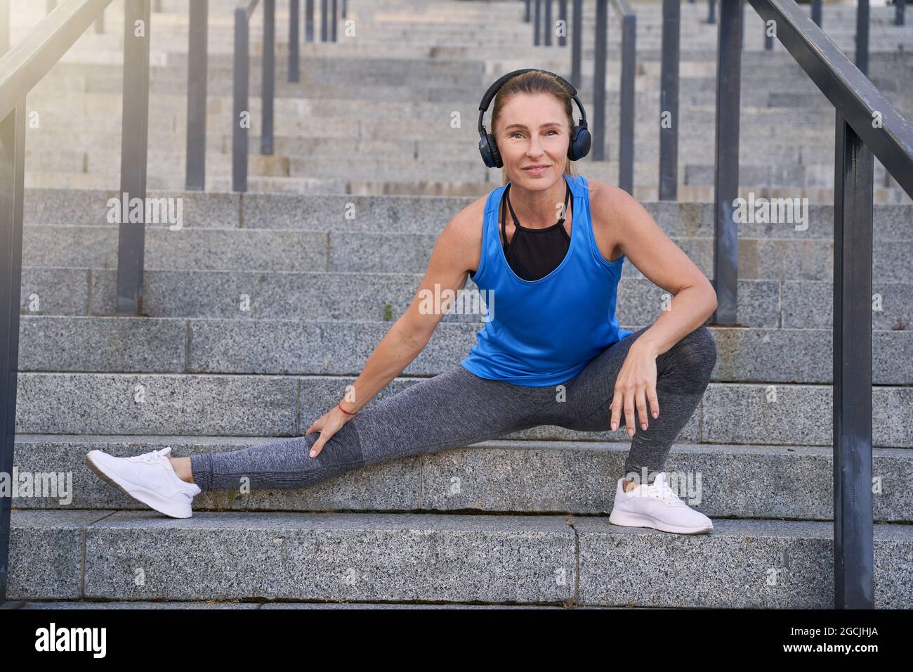 Fit middle aged woman in headphones looking at camera while stretching ...