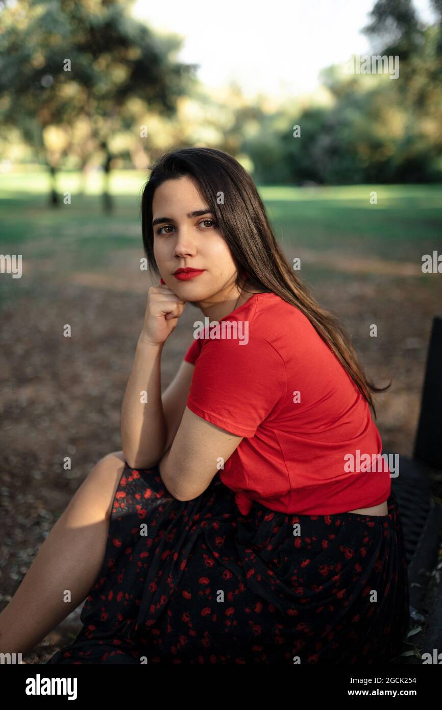 Side view of charming female chilling on bench and looking at camera while enjoying weekend in ...