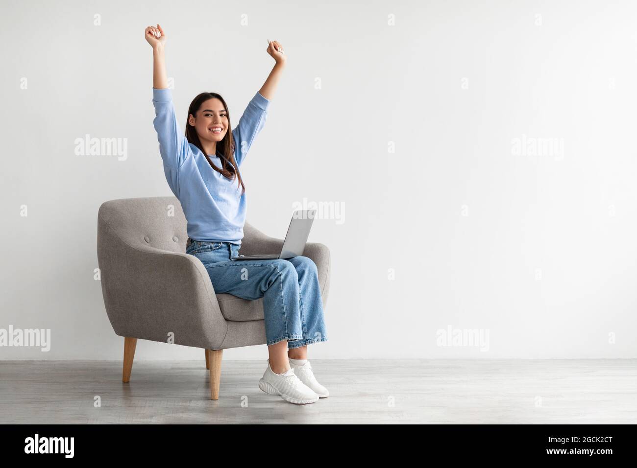 Ecstatic woman sitting in armchair with laptop, lifting hands up ...
