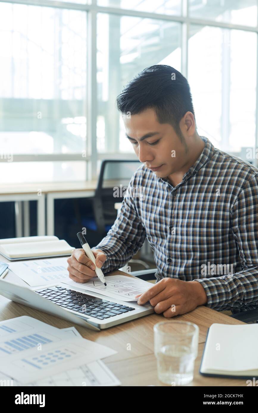 Serious young UI designer sitting at table with opened laptop and ...