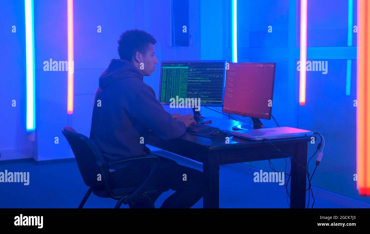 Young african man writing hacker programs in dark room with neon lights ...