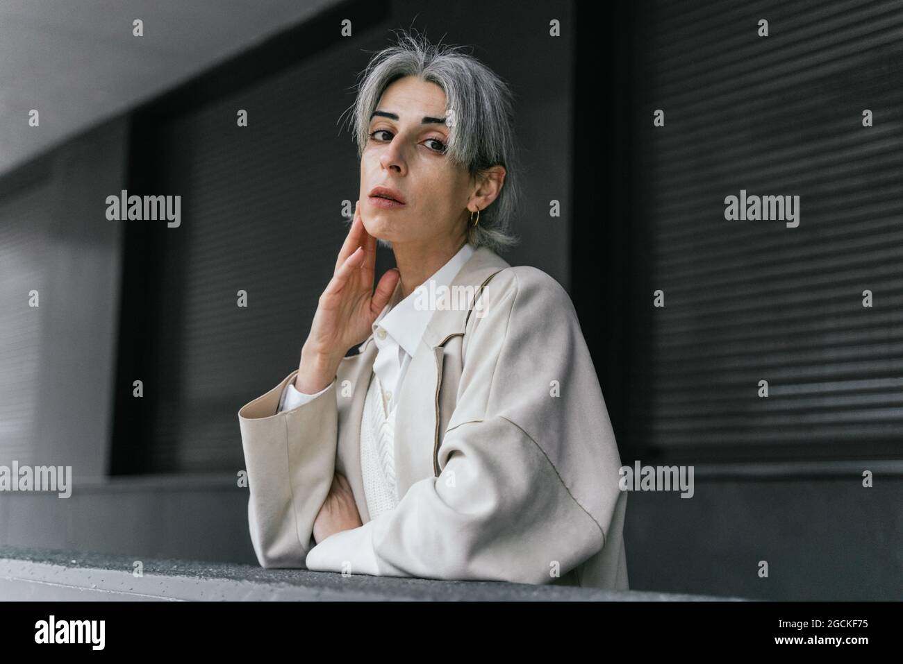 Serious transgender female in trendy coat leaning on gray fence while ...