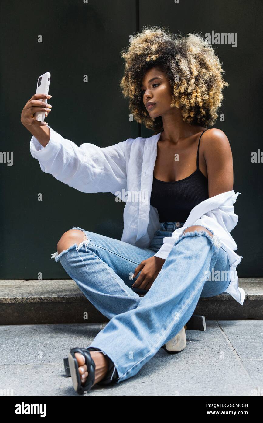 Full body of African American female in trendy casual outfit sitting on ...