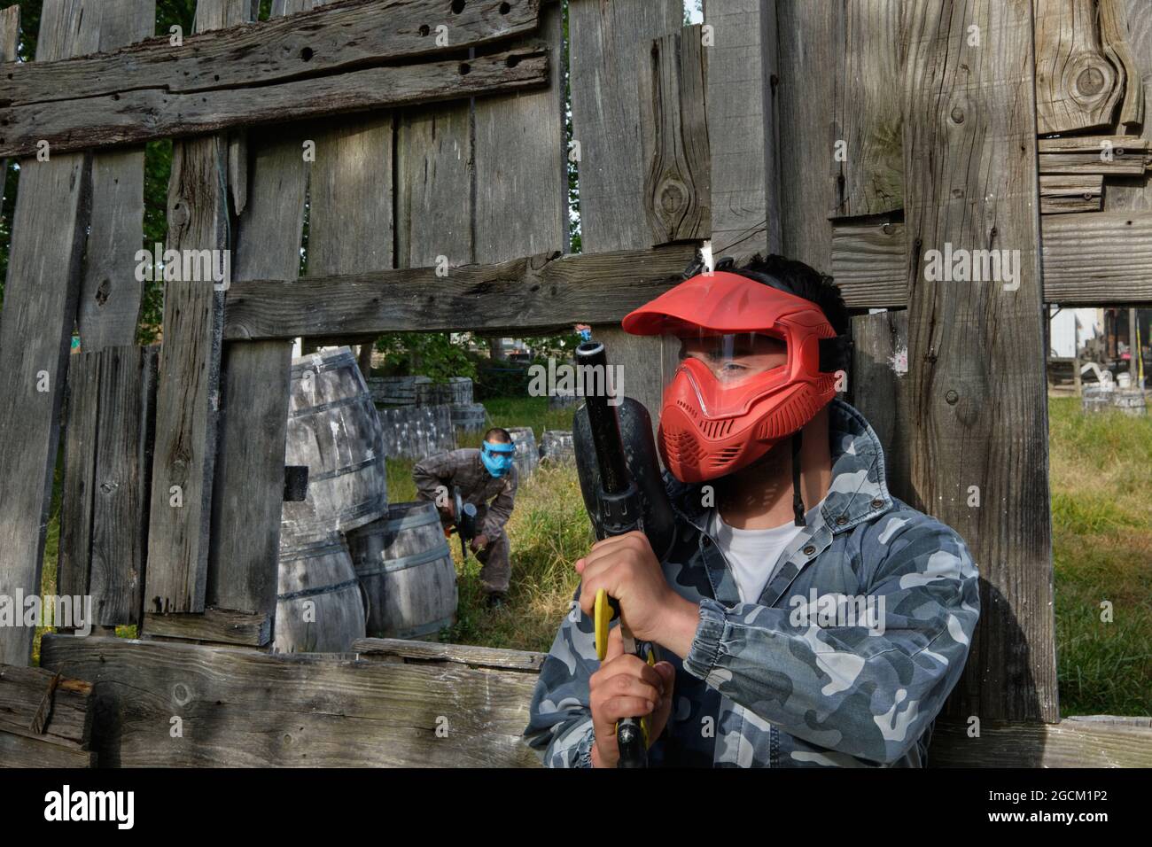 Male player in mask and with paintball gun standing behind wooden fence ...