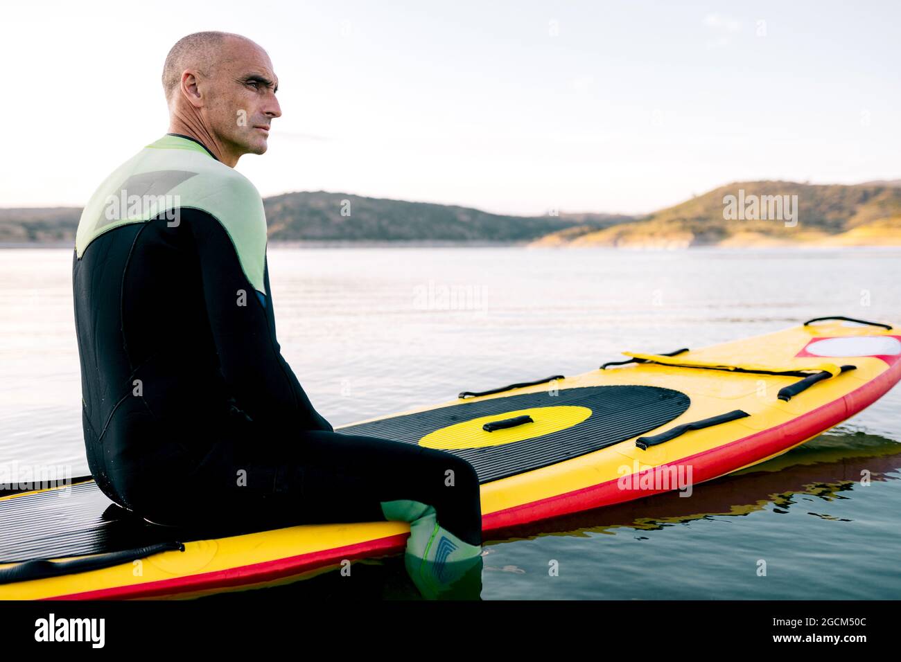 Side view of thoughtful adult male in wetsuit sitting on paddle board ...