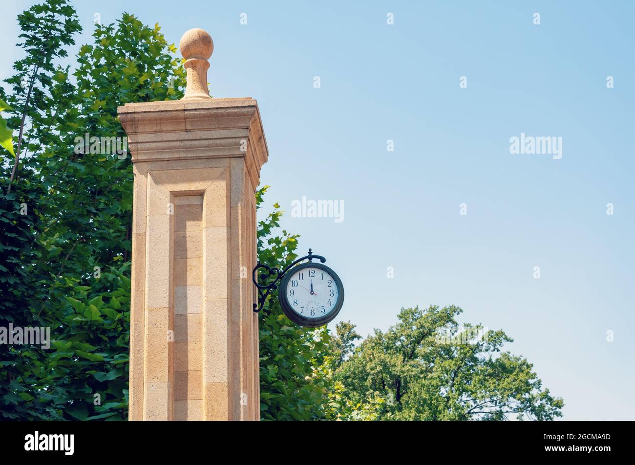 Street clock on a pole against blue sky and green foliage with copy ...