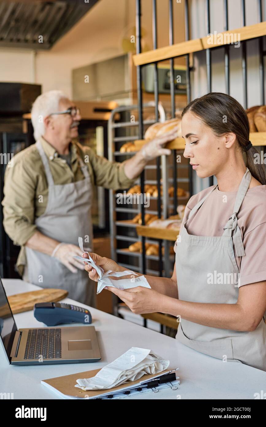 Busy female worker of bakery looking through receits while her mature ...