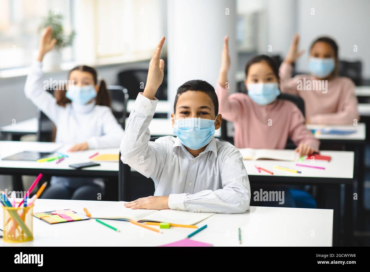 Schoolchildren raising hands at classroom, wearing protective medical ...