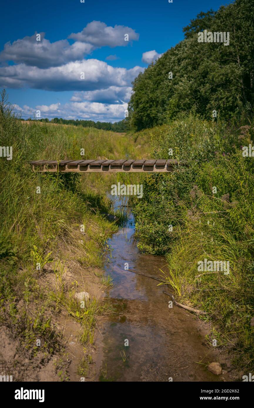 a small river with a wooden footbridge over it and sky clouds and ...