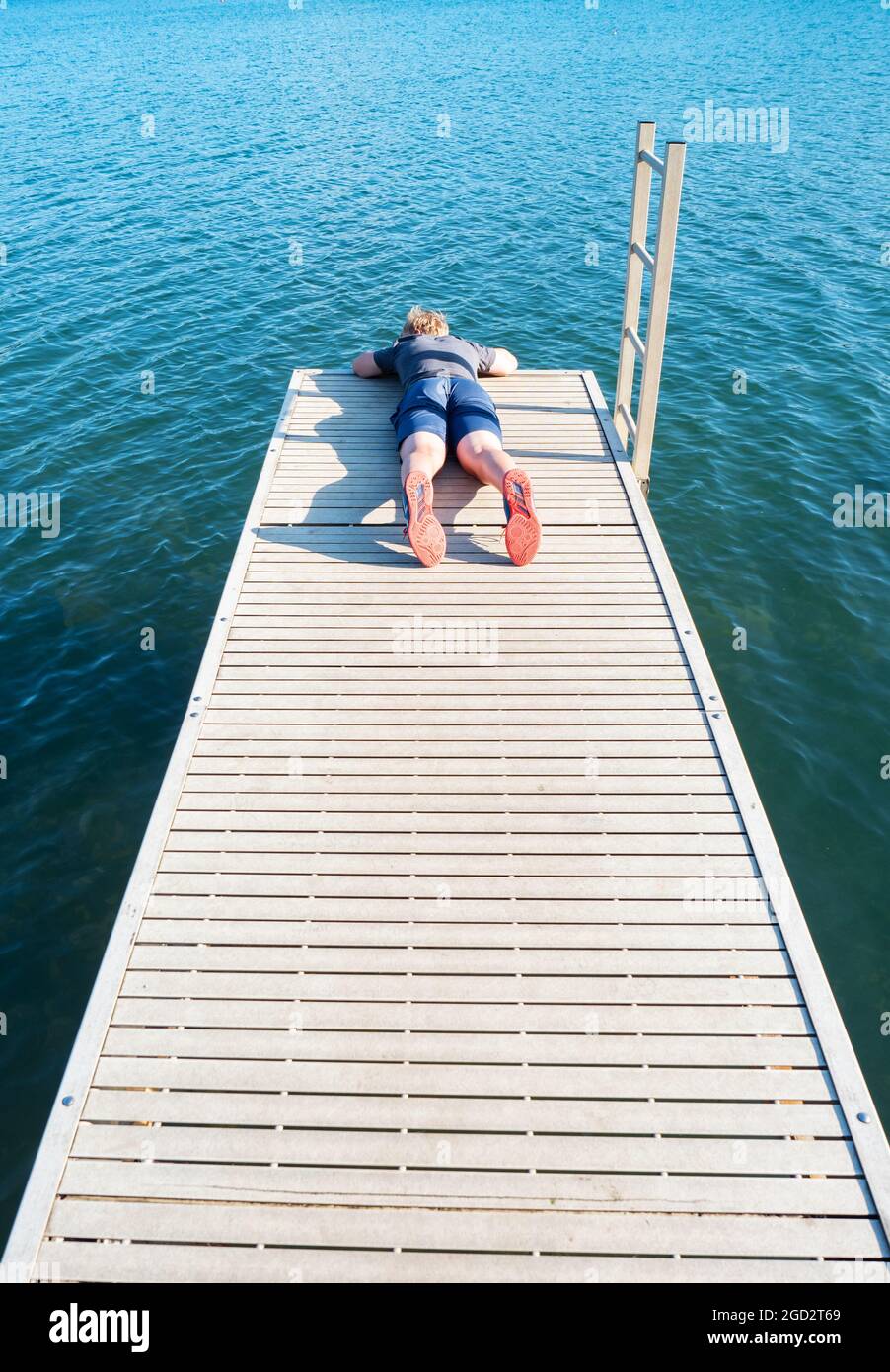 Young blond boy lying on a wooden pier over a lake watch fish in blue ...